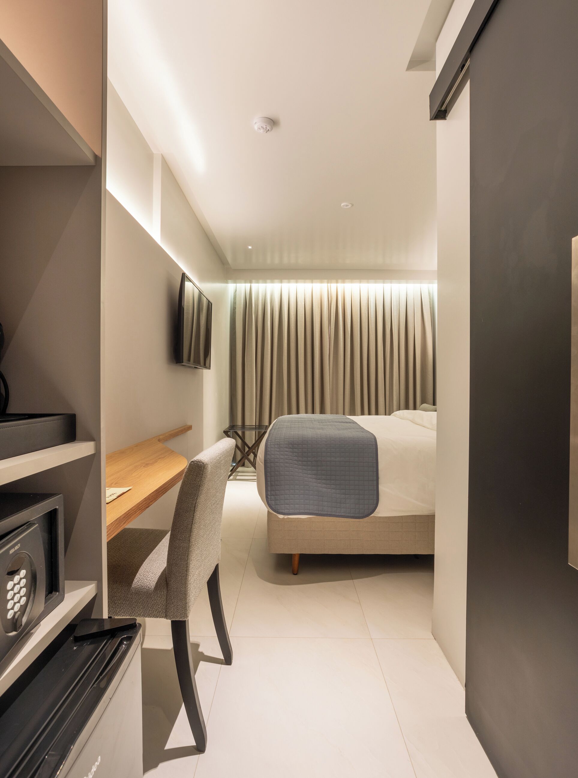 Quarto de hotel moderno com cama, escrivaninha, cadeira, televisão e minibar.