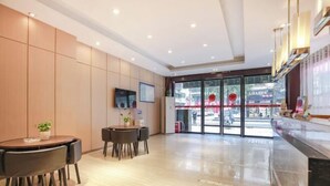 Um hall de hotel moderno com uma mesa de recepção de madeira, uma área de estar com uma mesa redonda e cadeiras, e uma grande porta de vidro que dá para a rua.