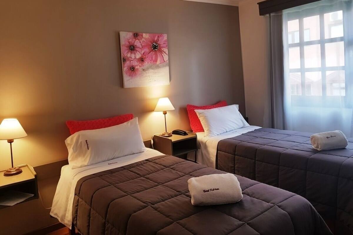 Quarto de hotel com duas camas, cada uma com travesseiro e cobertor. Há uma pintura na parede e uma janela com cortinas.