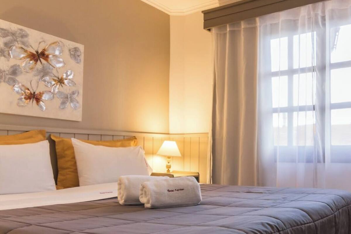 Quarto de hotel com uma cama grande, mesa de cabeceira com abajur e janela com cortinas.
