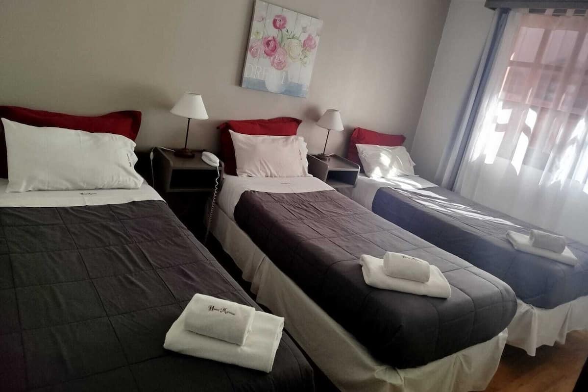 Quarto de hotel com três camas, cada uma com travesseiro e toalha dobrada. Há uma pintura na parede acima das camas e uma janela com cortinas.