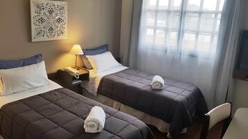 Quarto de hotel com duas camas, uma mesinha de cabeceira com luminária, uma obra de arte na parede e uma janela com cortinas translúcidas.