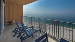 Varanda com duas cadeiras de descanso e uma mesa pequena, com vista para uma praia com guarda-sóis e pessoas.