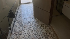 Um corredor estreito com ladrilhos estampados no chão, corrimão de metal e uma porta que dá para área externa.