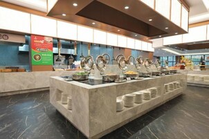 Buffet montado com várias estações de serviço, bancada de mármore e área de cozinha moderna ao fundo.