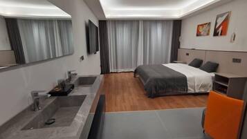 Quarto de hotel moderno com uma cama grande, escrivaninha embutida, espelho amplo, toucador com pia e chuveiro.