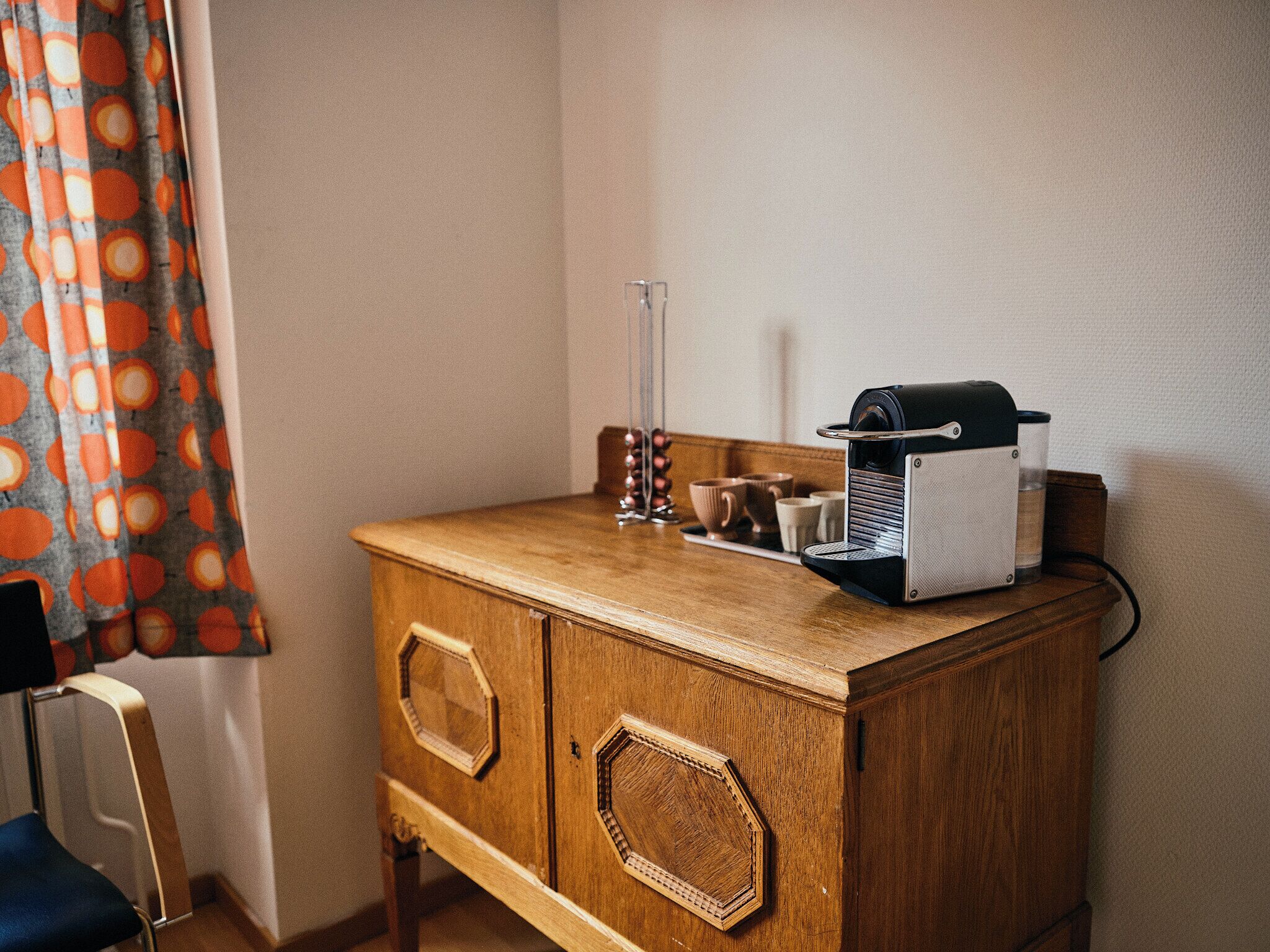 Uma cómoda de madeira com uma máquina de café e chávenas, uma cadeira e um cortínio com padrão de bolinhas.