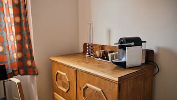 Uma cómoda de madeira com uma máquina de café e chávenas, uma cadeira e um cortínio com padrão de bolinhas.