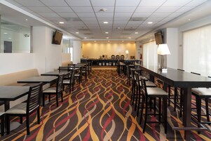 Uma área de refeições de hotel com várias mesas e cadeiras, um tapete estampado e um balcão com uma televisão.