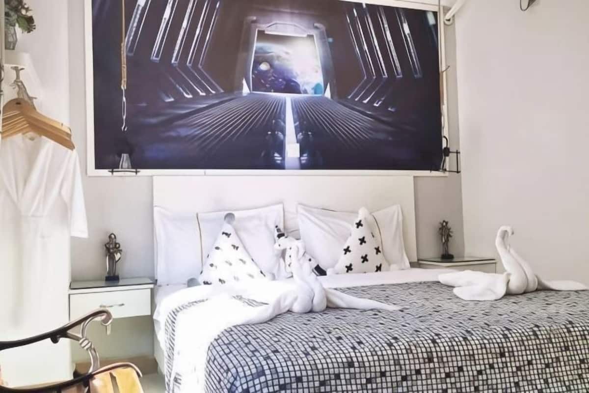 Quarto de hotel com uma cama, televisão na parede, mesa de cabeceira e uma cadeira.