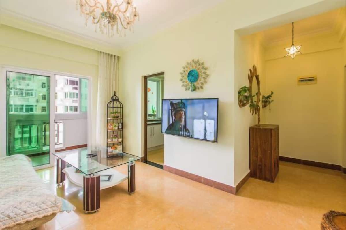 Uma sala de estar moderna com uma mesa de centro de vidro, uma TV de tela plana na parede, um lustre e vista para um edifício.