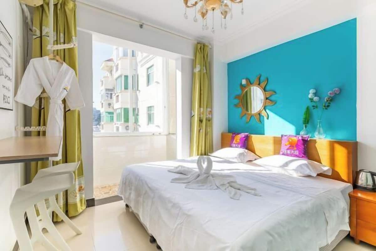 Quarto de hotel com cama, escrivaninha e cadeira. Há uma janela com cortinas, um lustre e um espelho em formato de sol na parede.