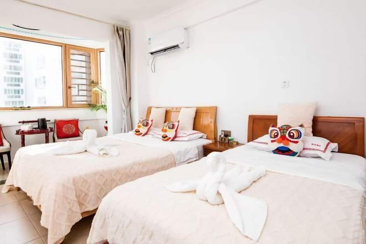Quarto de hotel com duas camas, uma janela, aparelho de ar condicionado e uma pequena mesa com um travesseiro vermelho.