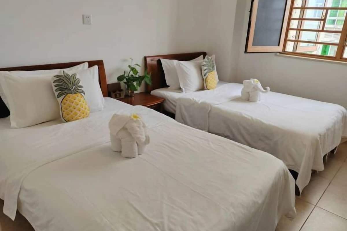 Quarto de hotel com duas camas de solteiro, cada uma com um travesseiro em formato de abacaxi e uma toalha modelada como elefante.