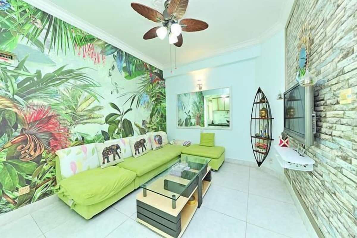 Uma sala de estar com um sofá verde, uma mesa de centro de vidro, um ventilador de teto, uma parede de pedra e uma TV à direita.
