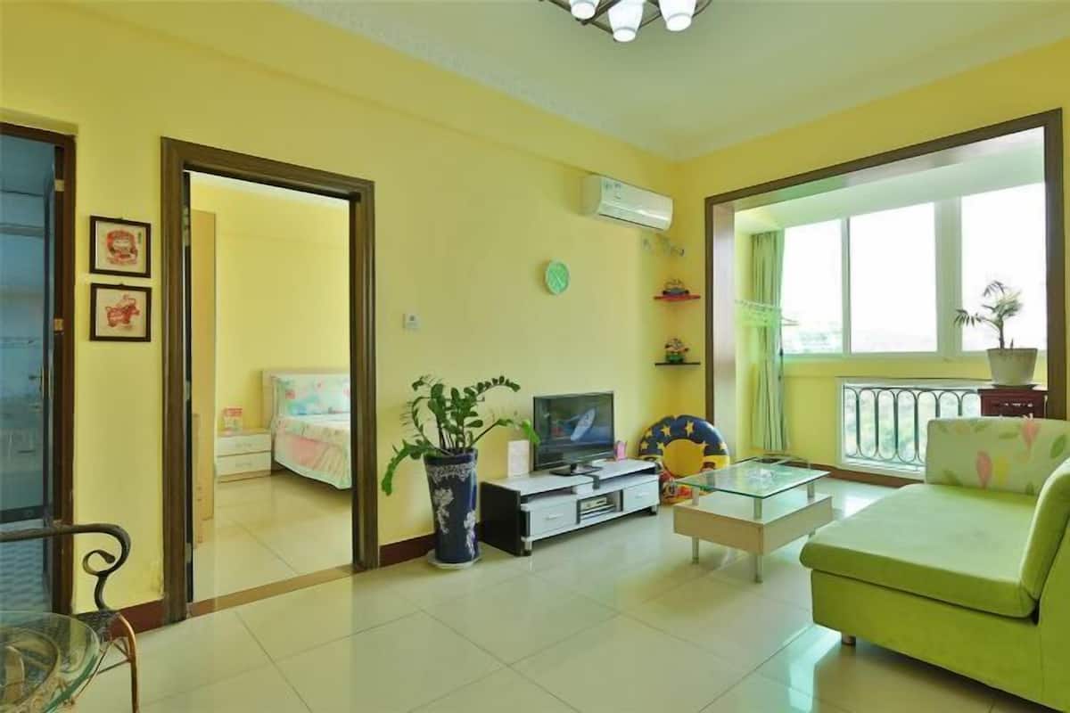 Sala de estar com sofá, televisão, mesa de centro de vidro, planta em vaso e varanda com guarda-corrimão.