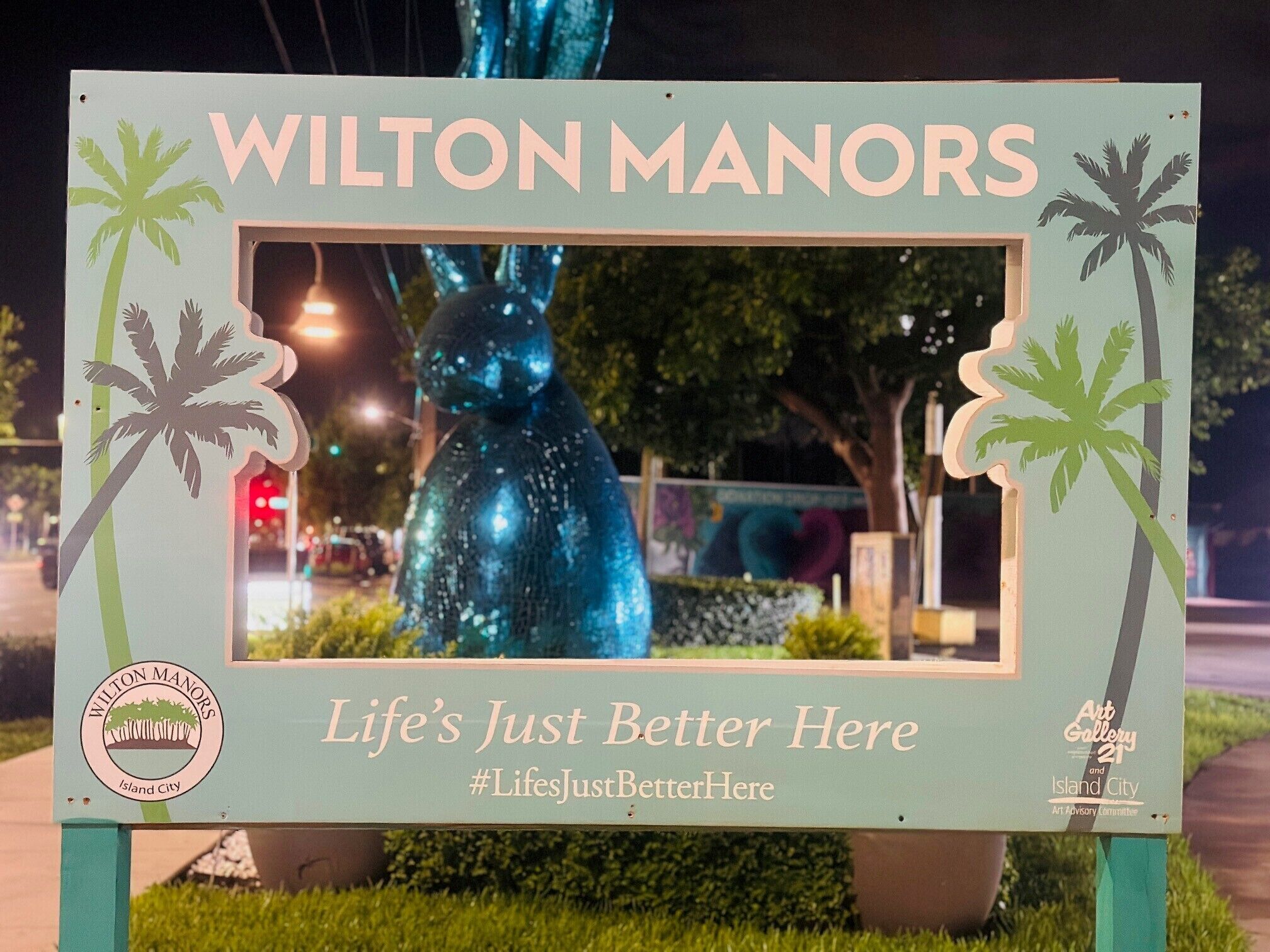 Um sinal para Wilton Manor, com moldura decorativa, silhuetas de palmeiras e os dizeres 'Wilton Manor' e 'A Vida é Melhor Aqui'.