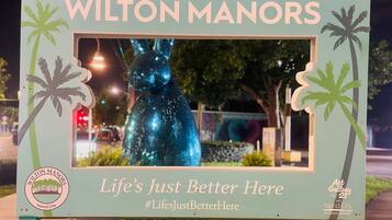 Um sinal para Wilton Manor, com moldura decorativa, silhuetas de palmeiras e os dizeres 'Wilton Manor' e 'A Vida é Melhor Aqui'.