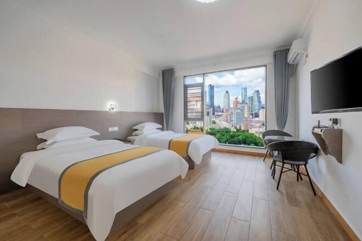 Quarto de hotel com duas camas, televisão, uma cadeira e uma janela grande com vista para a cidade.