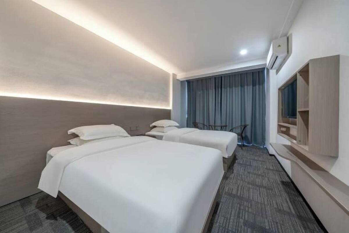 Quarto de hotel com duas camas, uma escrivaninha, uma cadeira, televisão e uma janela grande com cortinas.