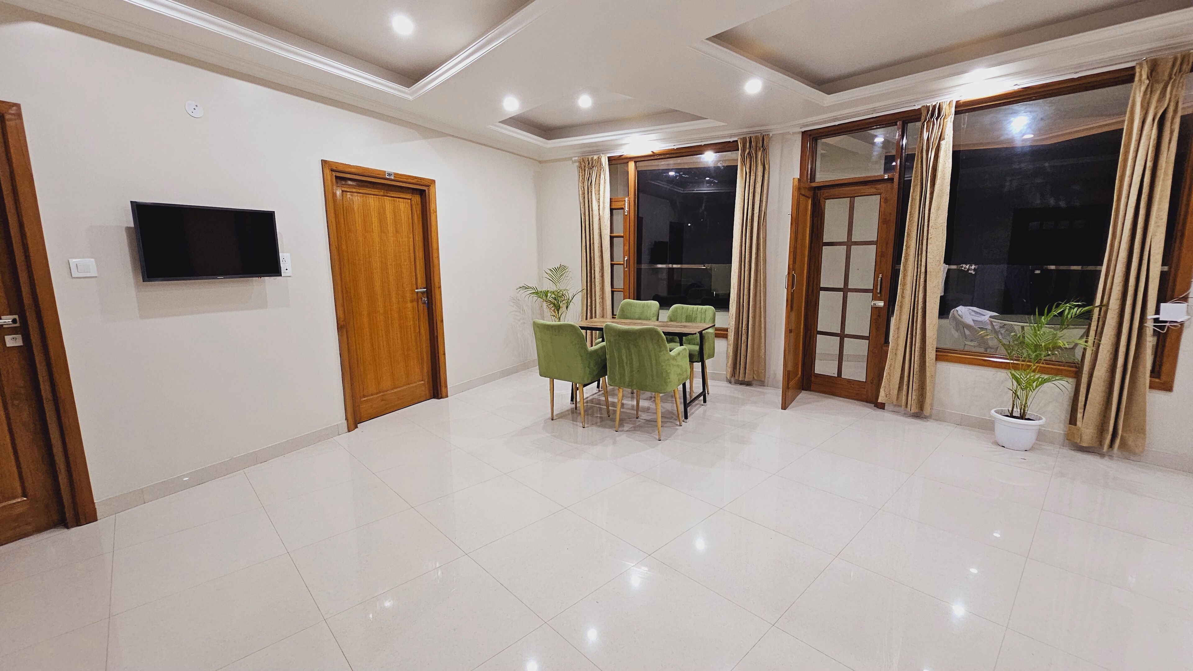 Um quarto amplo com uma mesa de jantar e cadeiras, televisão fixada na parede e grandes janelas com cortinas.