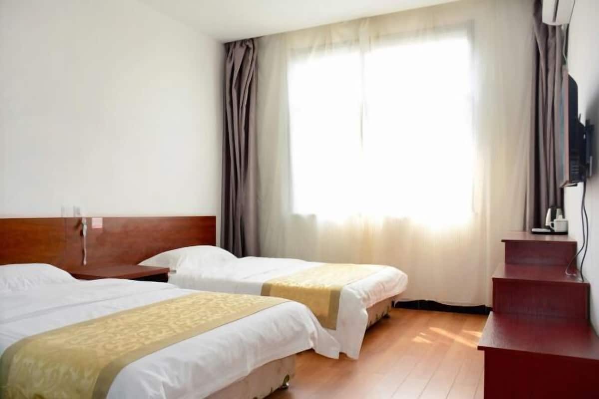 Quarto de hotel com duas camas, cabeceira de madeira, janela com cortinas e televisão na parede.