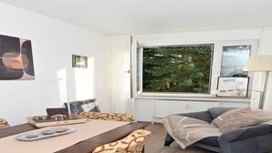 Uma sala de estar moderna com uma mesa de jantar de madeira, cadeiras estampadas, um sofá cinza e uma grande janela com vista para árvores.