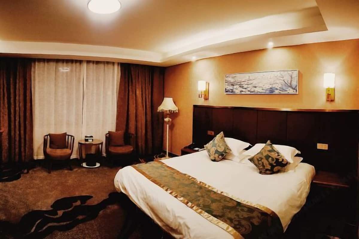 Quarto de hotel com uma cama grande, duas cadeiras, uma mesinha, um abajur e um quadro na parede.