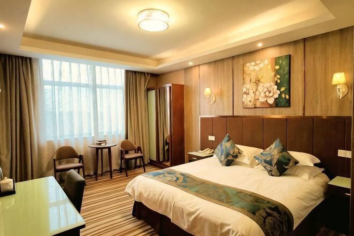 Quarto de hotel com uma cama grande, uma escrivaninha, duas cadeiras, um criado-mudo e um quadro na parede.