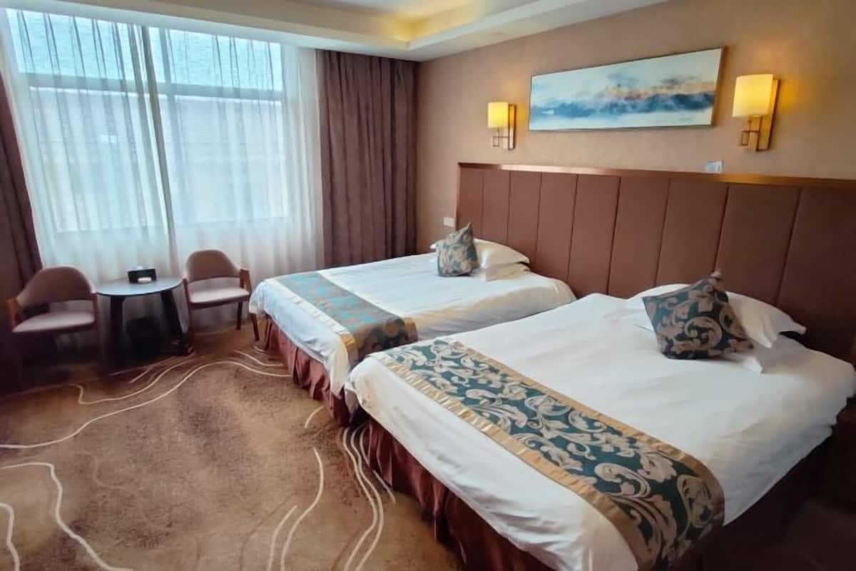 Quarto de hotel com duas camas, uma mesinha, cadeiras, uma janela grande com cortinas e luminárias fixadas na parede.
