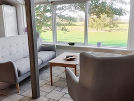 Quarto com sofá cinzento, uma mesinha com uma tigela e controles remotos, e janela com vista para uma área gramada.