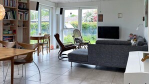 Uma sala de estar ampla com um sofá cinzento, uma mesa de jantar de madeira com cadeiras, uma estante de livros e uma televisão na parede.