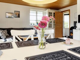 Uma área de refeições com uma mesa posta para três, apresentando jogos americanos pretos com texto, um vaso com flores cor-de-rosa e uma vela sobre a mesa.
