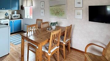 Uma sala de jantar com uma mesa e cadeiras de madeira, uma cozinha com armários azuis, um micro-ondas e uma televisão na parede.
