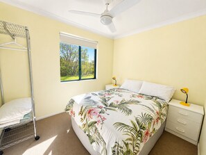 Quarto com cama, janela, ventoinha no teto, mesa de cabeceira, abajur e um suporte para roupas.