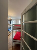 Quarto com beliche, um lustre, uma porta com painéis de vidro jateado e vista para outro quarto com cama e janela.