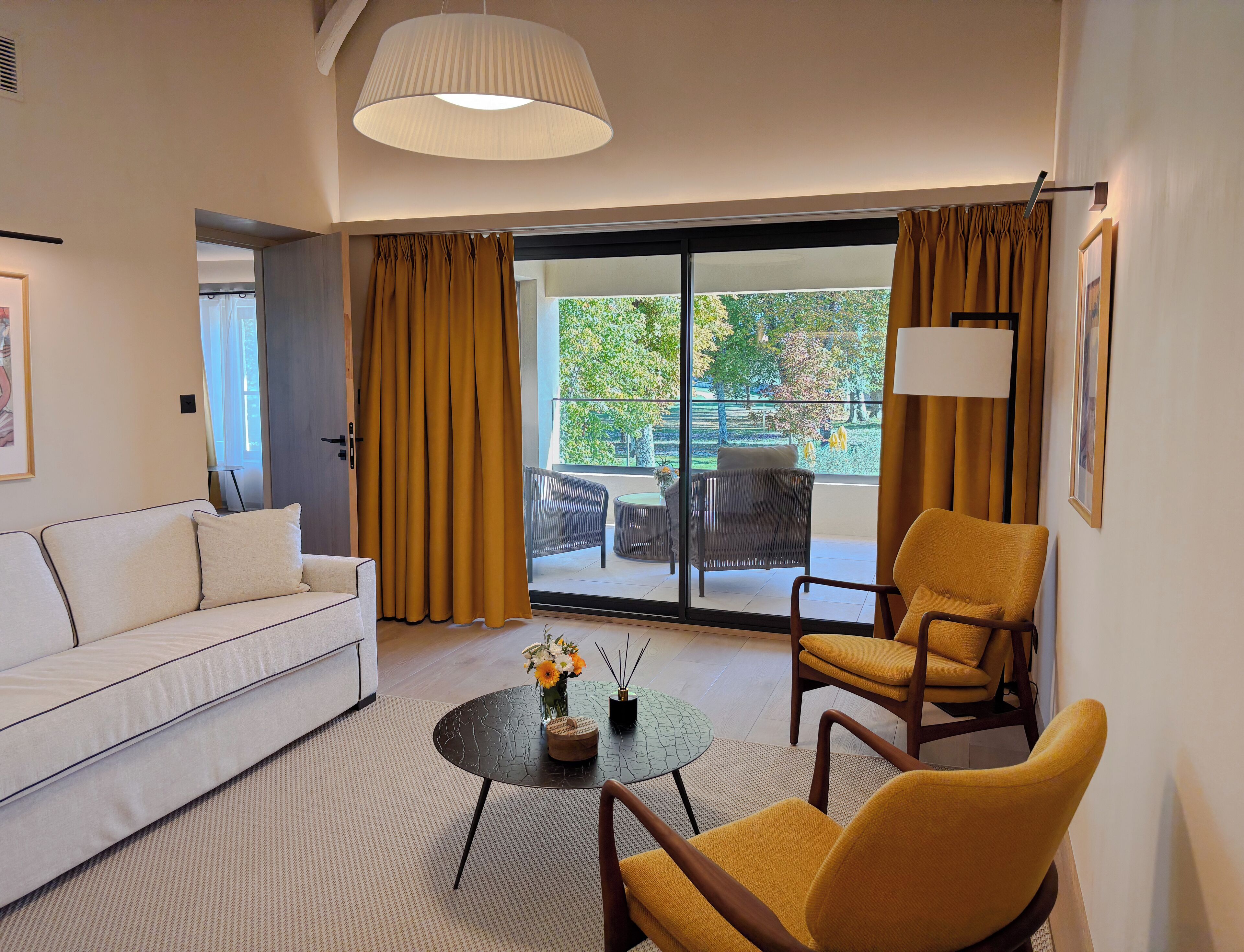Quarto de hotel moderno com sofá branco, poltrona amarela, mesa redonda pequena com um vaso de flores e uma grande porta de vidro deslizante que dá para uma área externa com vista para o verde.