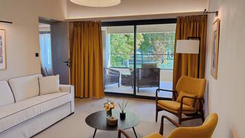 Quarto de hotel moderno com sofá branco, poltrona amarela, mesa redonda pequena com um vaso de flores e uma grande porta de vidro deslizante que dá para uma área externa com vista para o verde.
