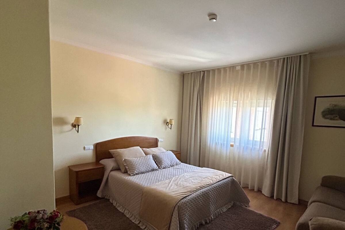 Quarto de hotel com cama, mesa de cabeceira, cadeira, mesa com um vaso de flores e janela com cortinas.