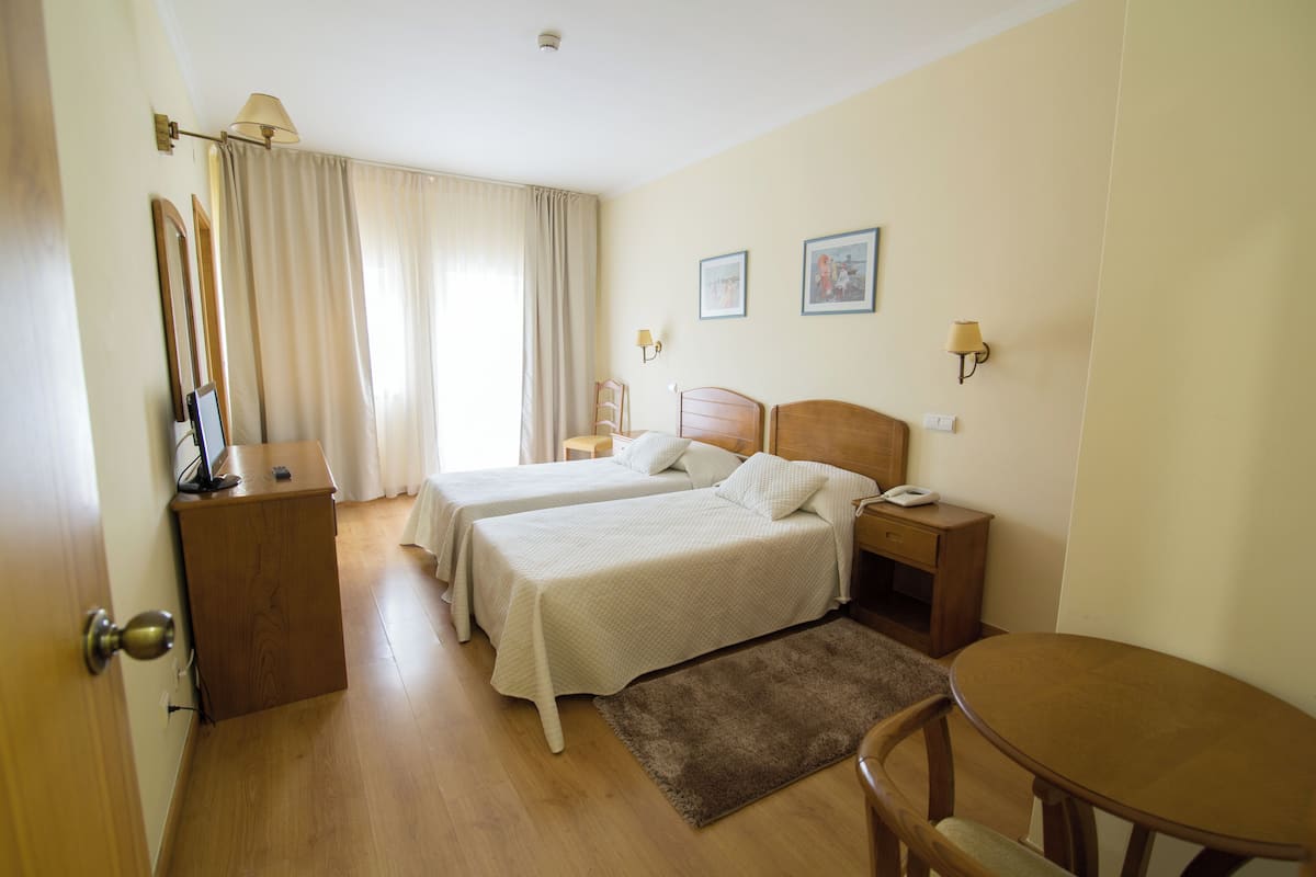 Quarto de hotel com duas camas, uma escrivaninha, uma cadeira, uma mesa e um abajur.