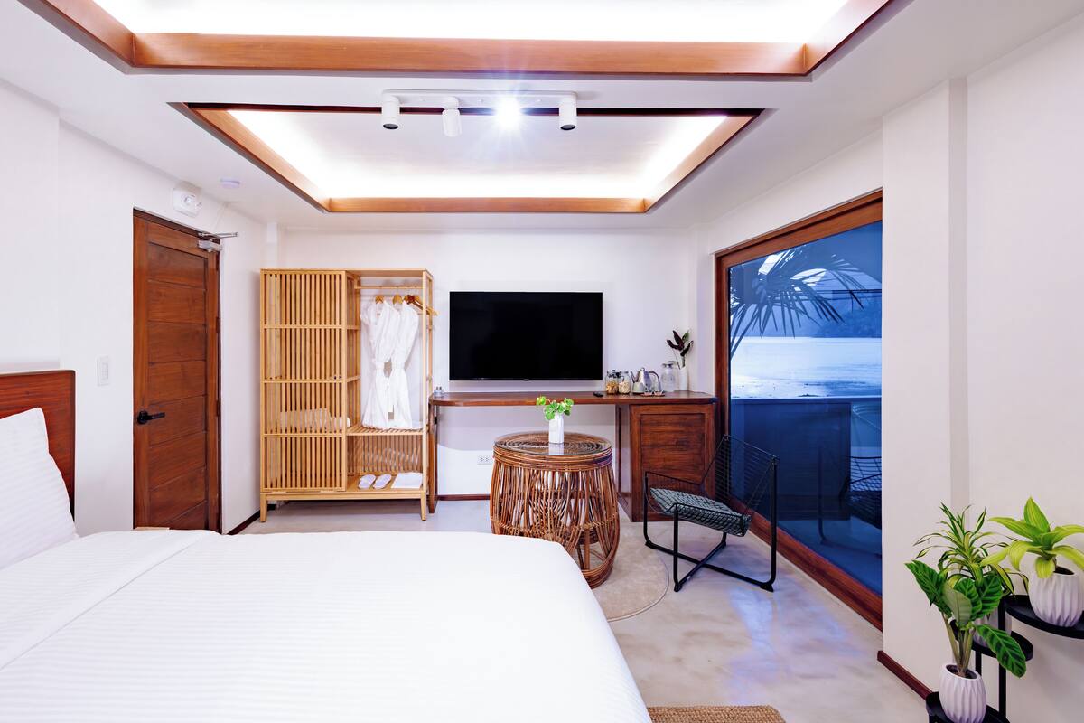 Quarto de hotel moderno com uma cama grande, televisão, guarda-roupa, uma mesinha com uma planta e varanda com vista.