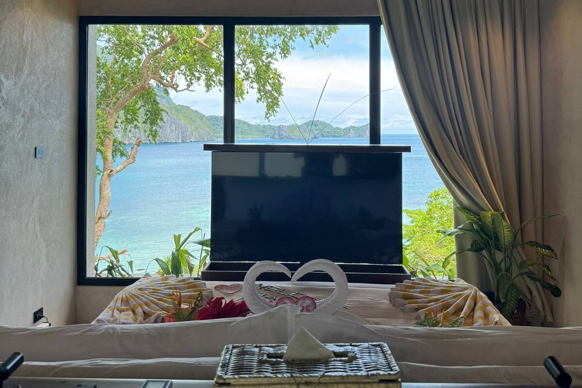 Quarto com uma grande janela que oferece vista para o mar e montanhas. Há uma televisão de tela plana em um suporte, uma pequena mesa com uma cesta e uma decoração em formato de coração, e um controle remoto sobre a mesa.