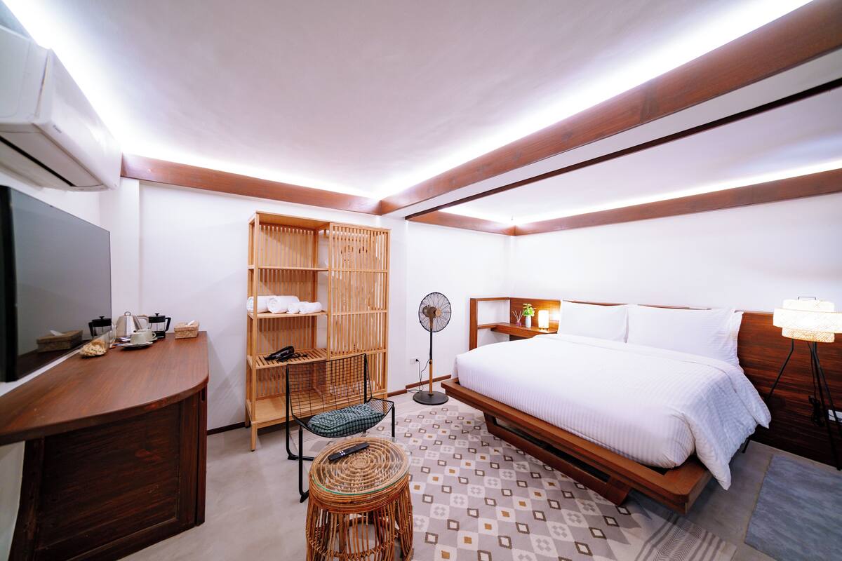 Quarto de hotel com cama, mesa de cabeceira, televisão, guarda-roupa e ventoinha.