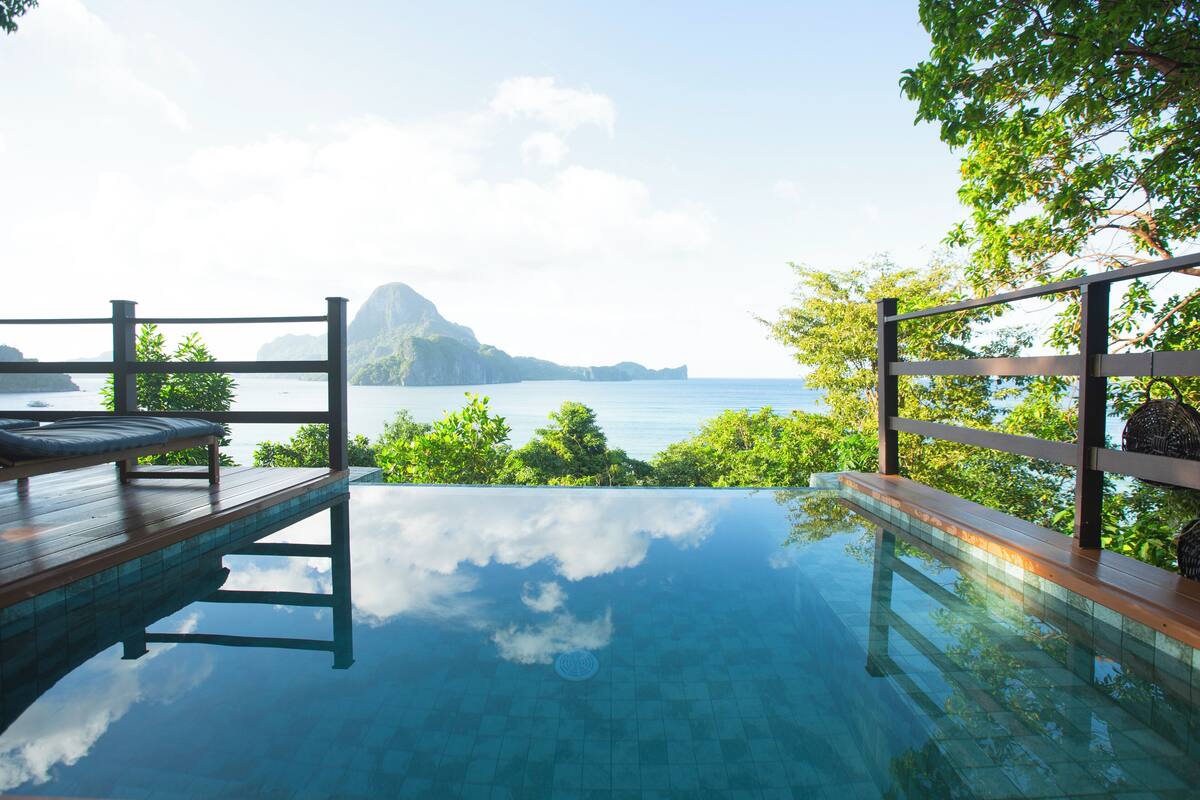 Uma piscina infinita com deck e guarda-corpo de madeira, cercada por vegetação exuberante e vista para o mar e montanhas ao longe.