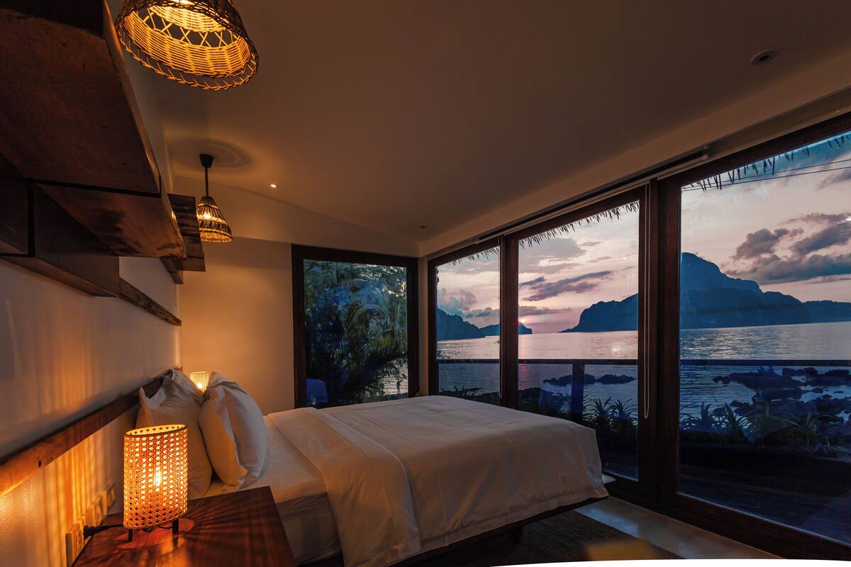 Quarto com uma grande janela que dá para um corpo d'água e montanhas, uma cama com lençóis brancos e uma luminária de parede texturizada.
