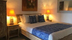 Cama bem arrumada com colcha florida azul, duas luminárias de cabeceira, mesa de cabeceira de madeira e um quadro na parede.