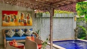 Uma área de estar coberta ao ar livre, com piscina, um muro de pedra e uma pintura.