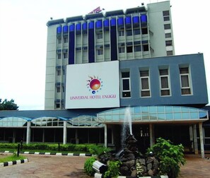 Um hotel com uma grande placa indicando "UNIVERSAL HOTEL ENUGU" e um recurso de água em frente.