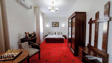 Quarto de hotel com tapete vermelho, cama com roupa de cama branca, guarda-roupa de madeira, espelho, um tabuleiro de xadrez e escrivaninha com abajur.