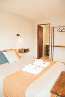 Cama bem arrumada com almofadas e manta, mesa de cabeceira de madeira, luminária na parede e porta que dá para um banheiro.
